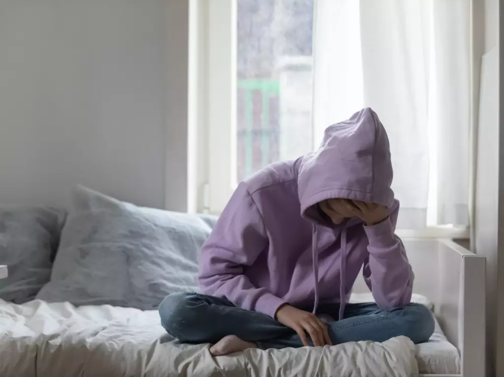 Olycklig ung 12-års flicka som sitter på bekväm säng och känner sig utmattad eller deprimerad ensam hemma. Stressat tonårsbarn som lider av huvudvärk eller har psykiska problem.