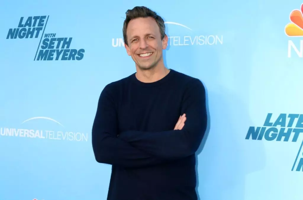 Seth Meyers in piedi con indosso un maglione blu con le braccia incrociate.