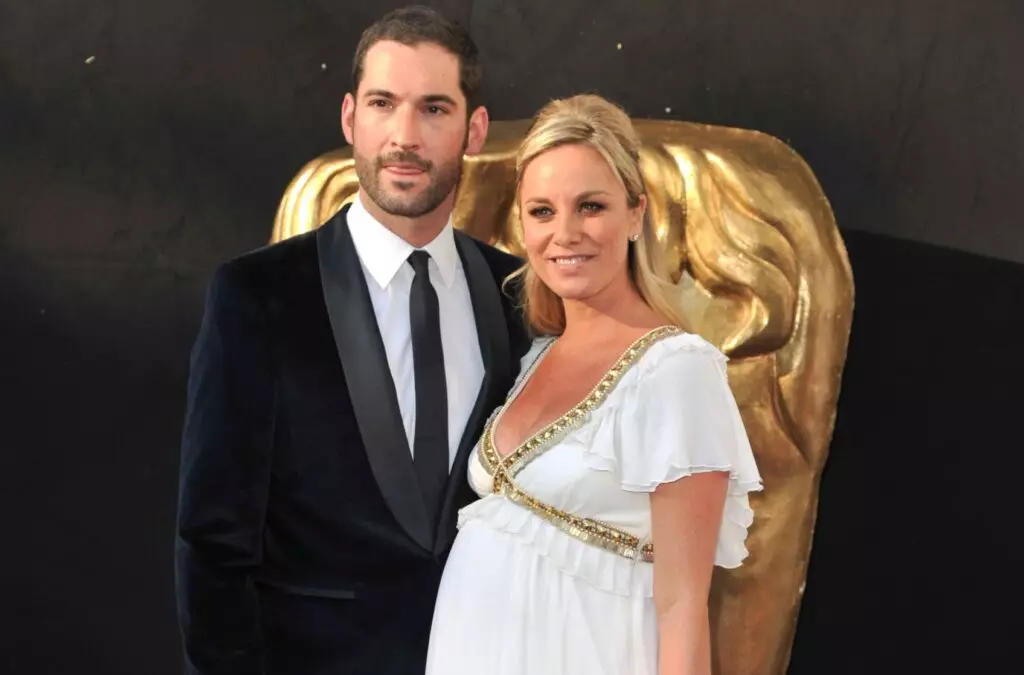 Tom Ellis i Tamzin Outhwaite na czerwonym dywanie. Tom ma na sobie czarny garnitur. Tamzin jest w ciąży w biało-złotej sukience.