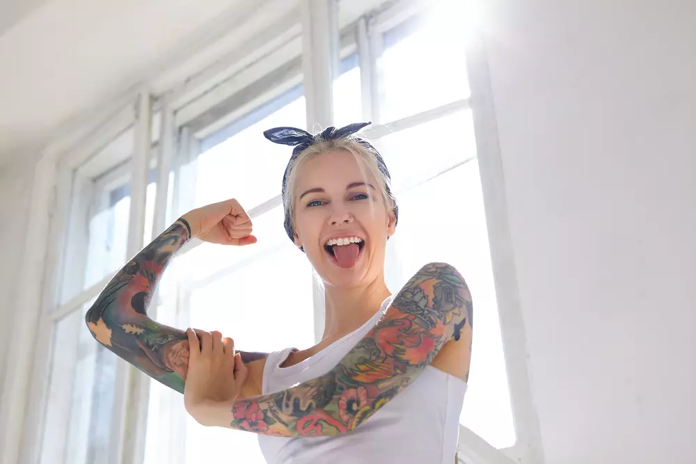 Photo ensoleillée d'une jeune jolie femme blonde joyeuse avec des tatouages montrant la langue joyeusement à la caméra tout en démontrant ses forts biceps, posant devant une grande fenêtre