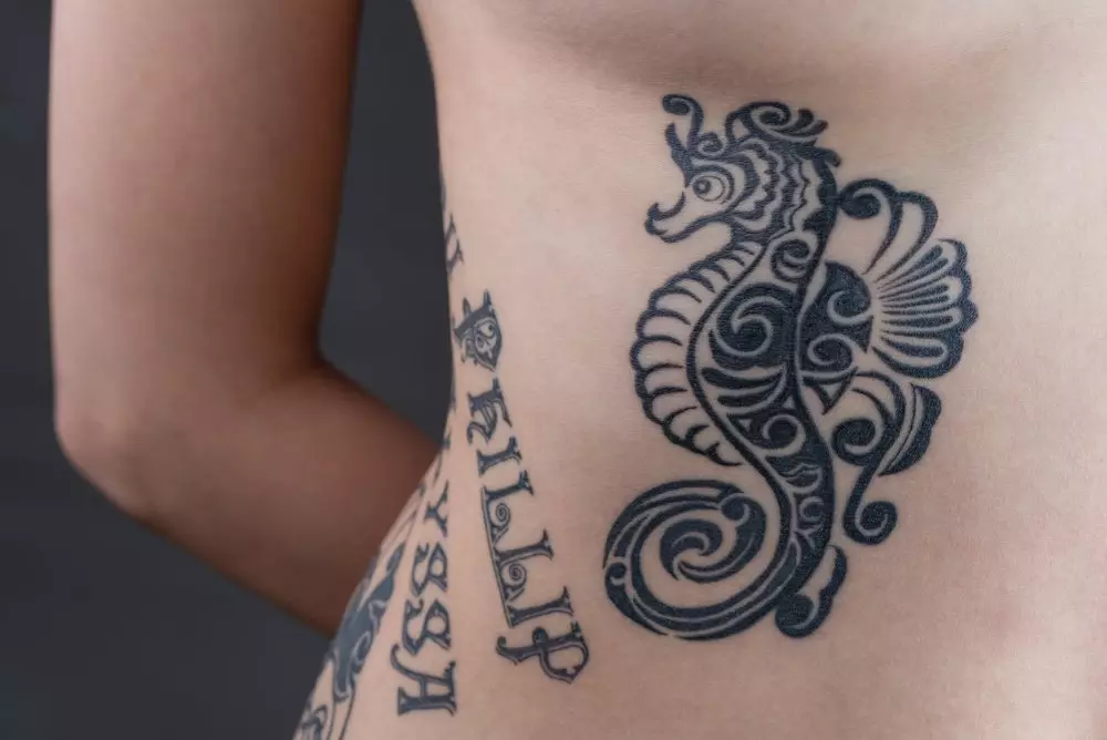 Un tatouage d'un hippocampe de style tribal noir avec des noms d'enfants s'enroulant autour des côtes d'une femme.